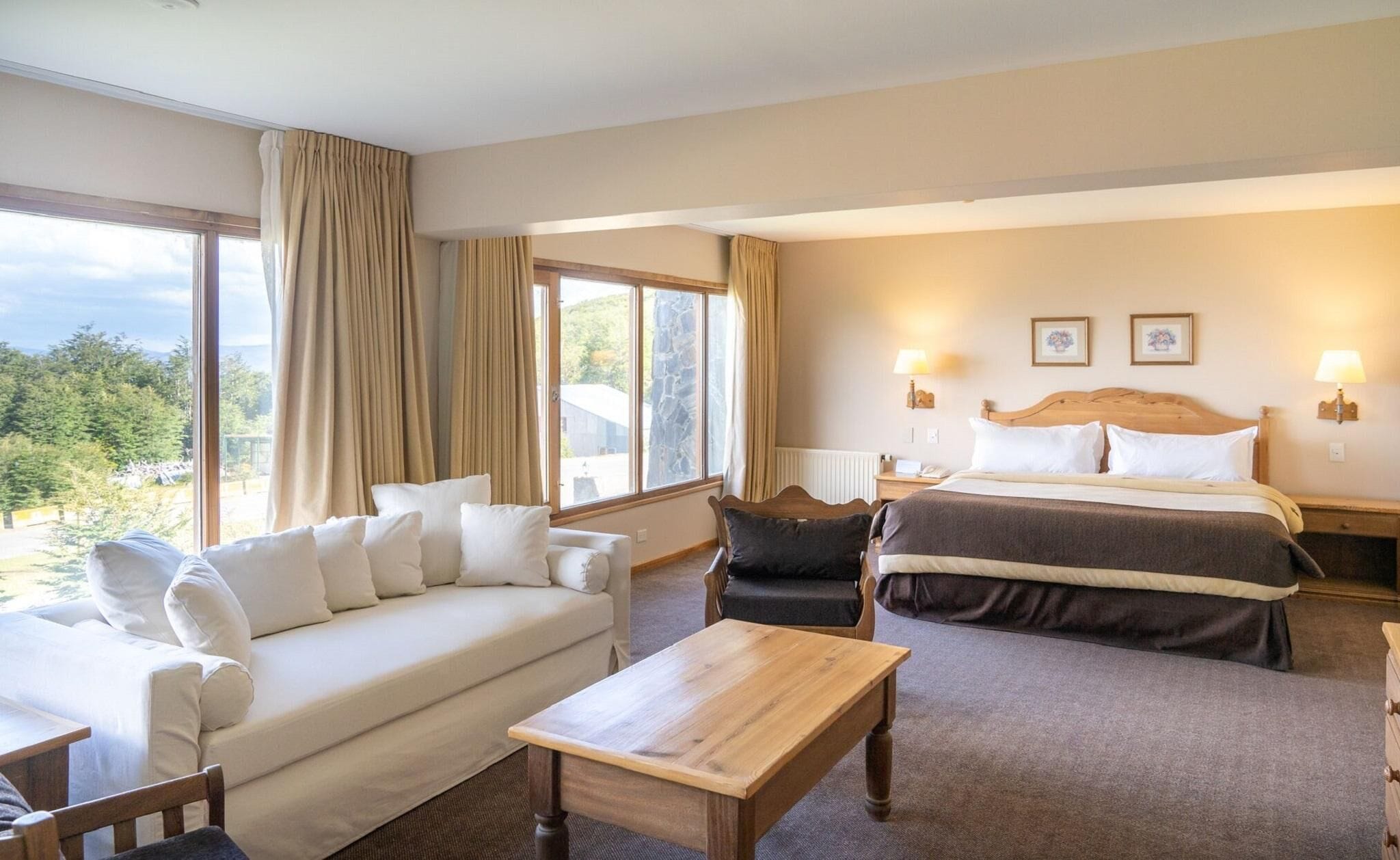 Quarto de hotel com sofá, cama, mesa de madeira e janelas grandes com cortinas.
