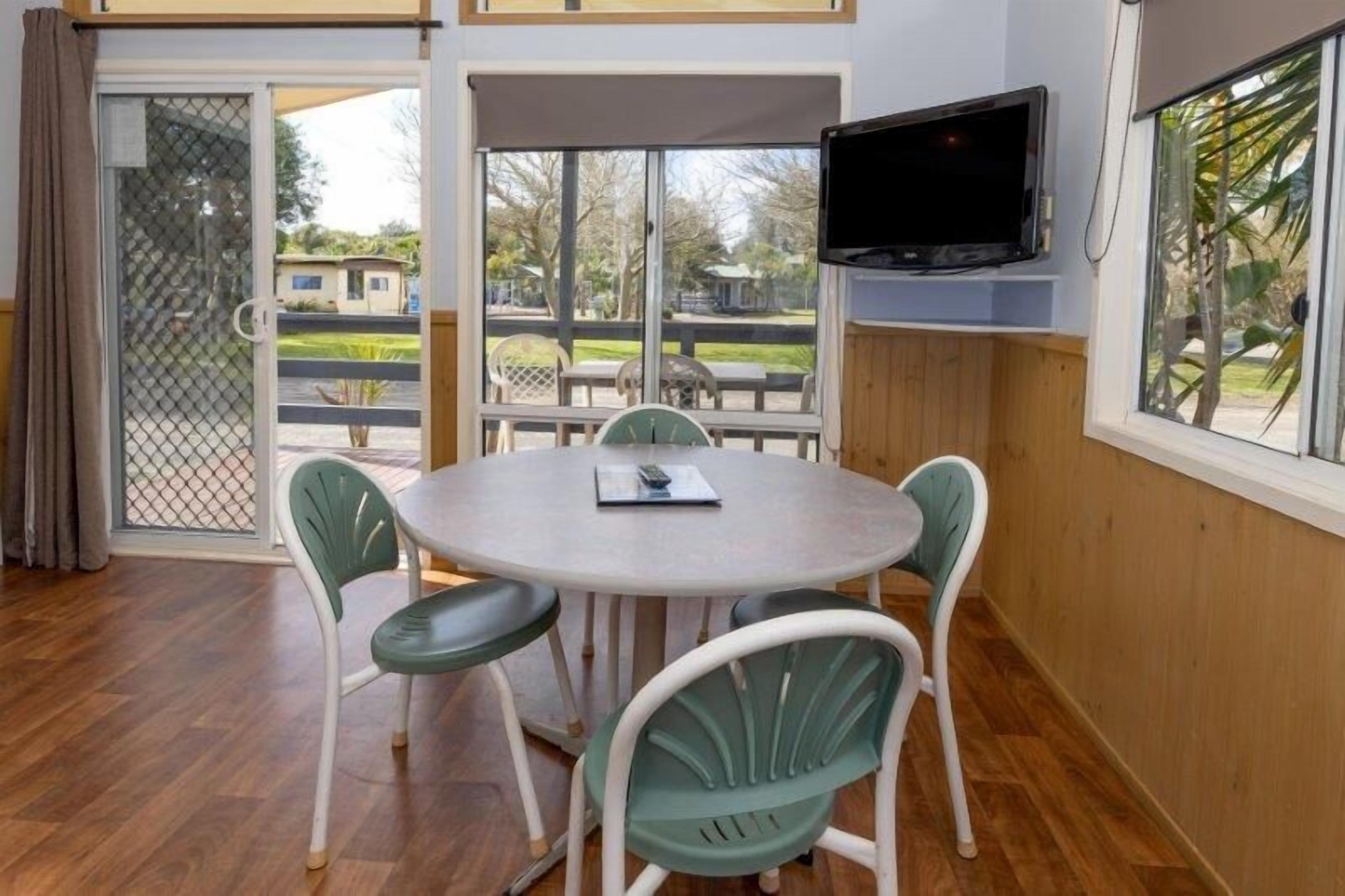 Quarto com mesa redonda e quatro cadeiras, televisão na parede e porta de vidro deslizante para área externa.