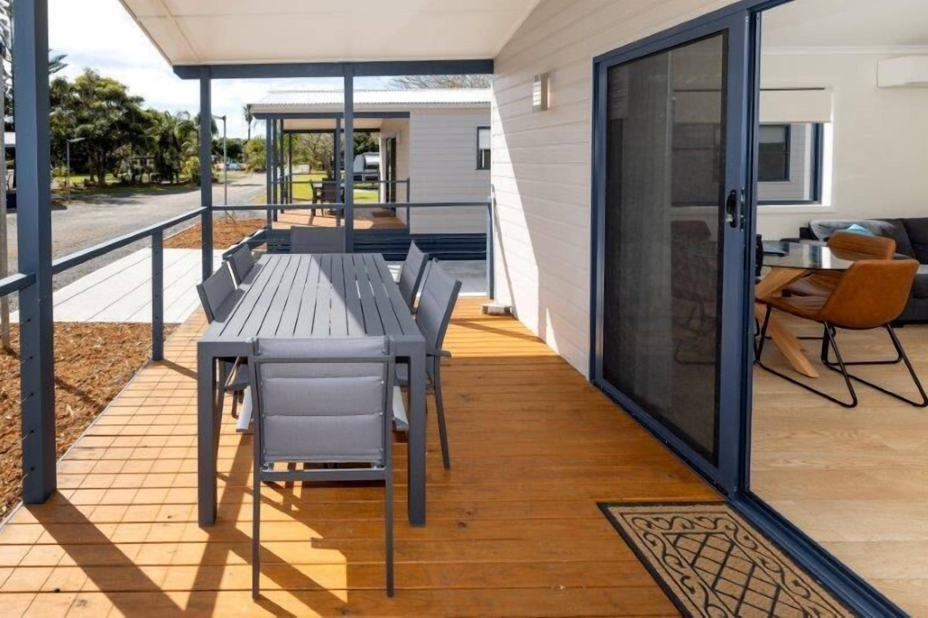Uma área externa coberta com um deck de madeira, uma mesa com cadeiras e uma porta de vidro deslizante que dá acesso a um espaço interno com um sofá e uma mesa de centro.