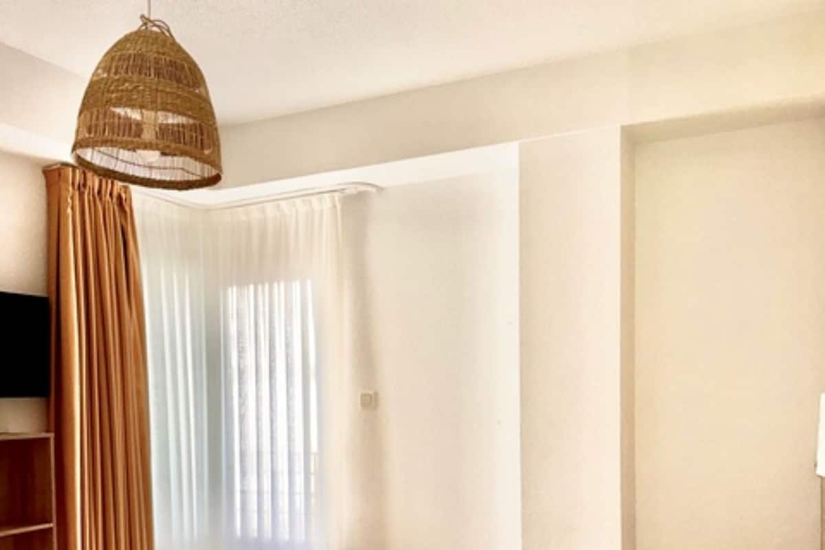 Quarto de hotel com cama, cortinas, televisão e luminária pendente.