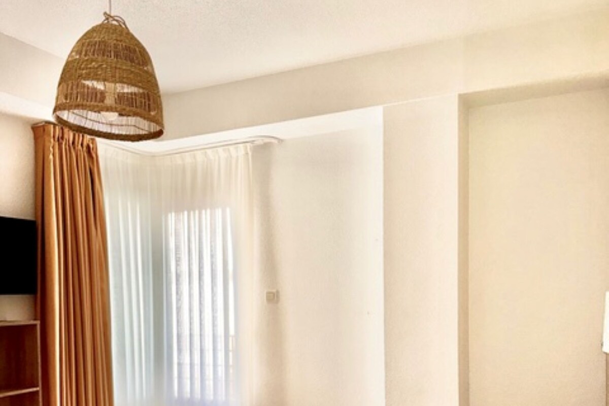 Quarto de hotel com cama, cortinas, televisão e luminária pendente.