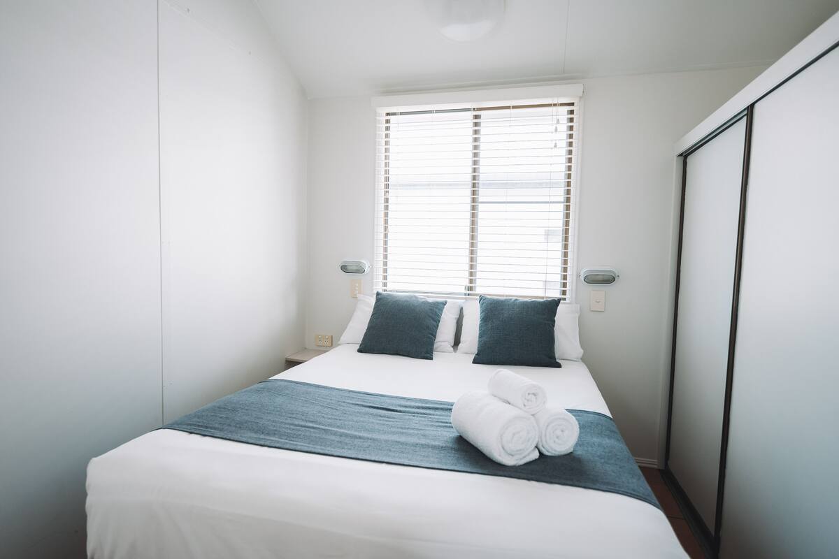 Uma cama bem arrumada com roupa de cama branca e azul-turquesa, uma janela com persianas e um guarda-roupa à direita.
