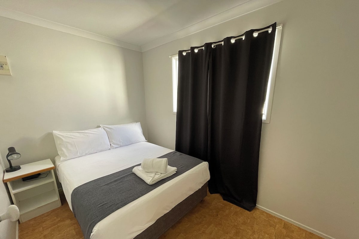 Quarto de hotel com uma cama, mesa de cabeceira, abajur, janela com cortinas e piso coberto de carpete.