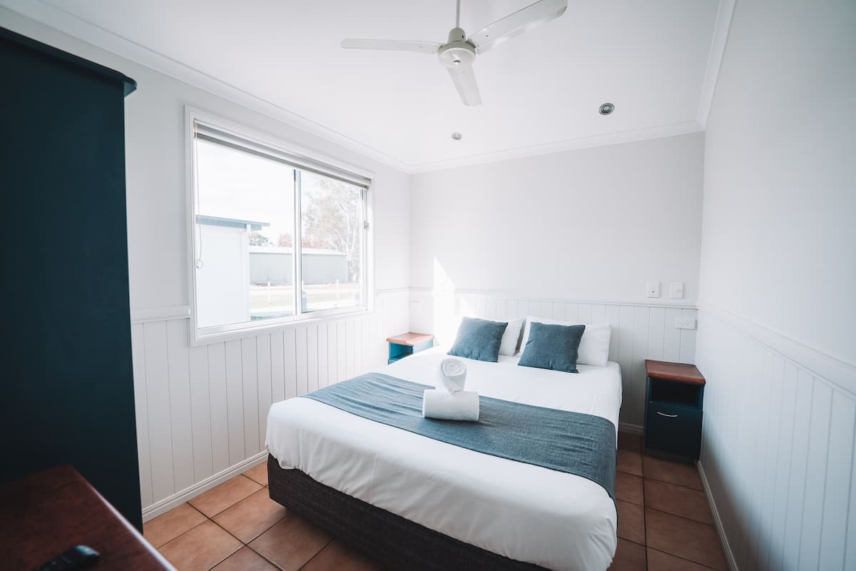 Quarto com cama, mesa de cabeceira, janela e ventoinha no teto.