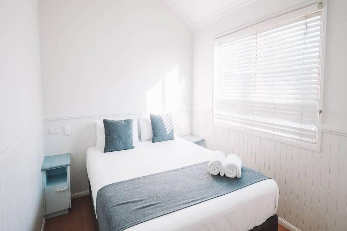 Quarto com cama, duas almofadas, mesa de cabeceira, janela com persianas e toalha sobre a cama.