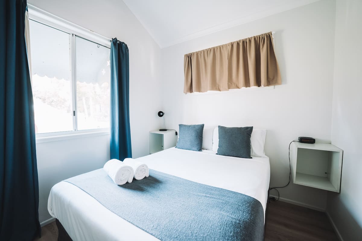 Uma cama bem arrumada com roupa de cama branca e um cobertor azul, uma mesa de cabeceira com abajur e uma janela com cortinas.