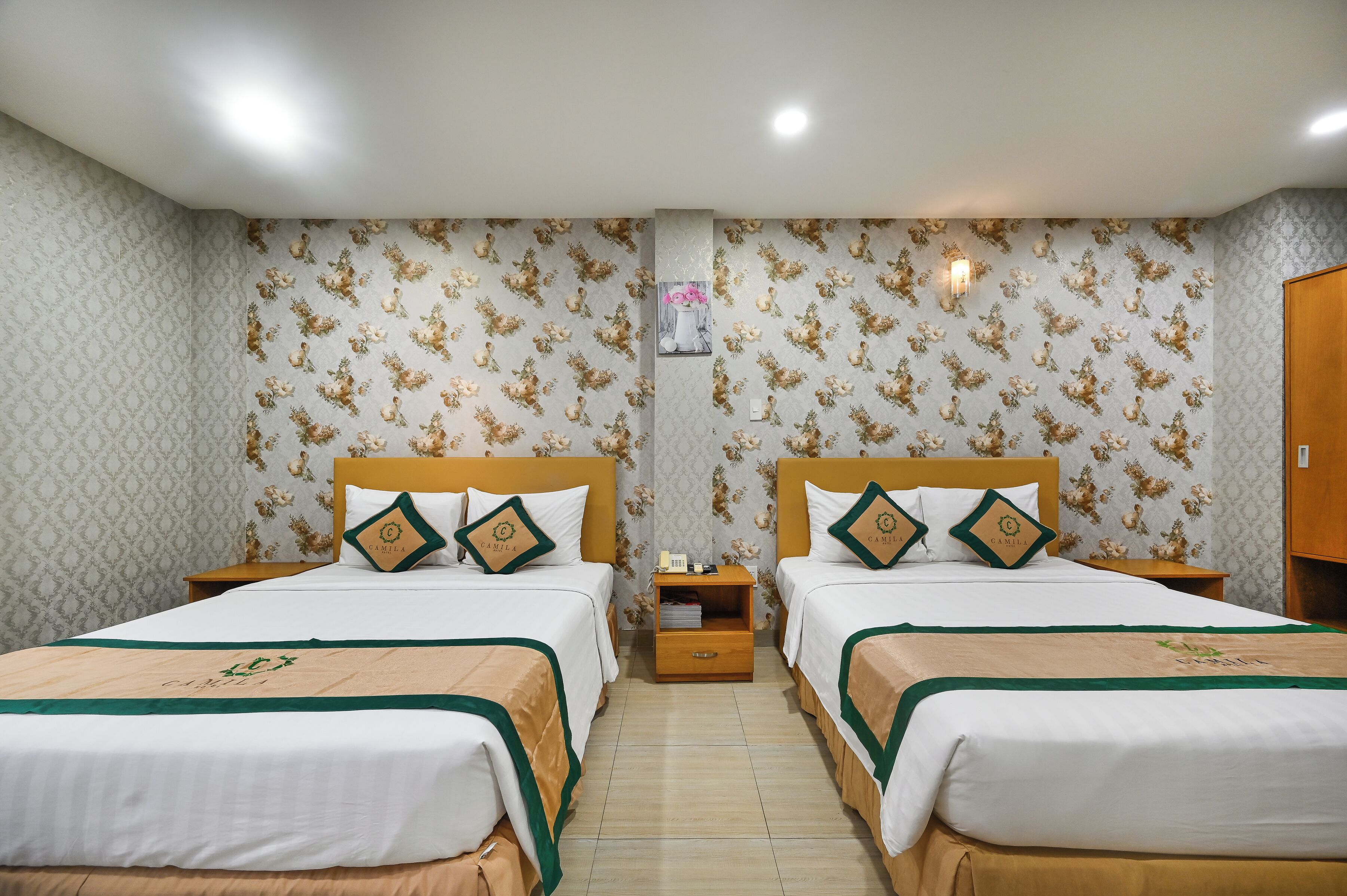 Quarto de hotel com duas camas, cada uma com cabeceiras e almofadas combinando, uma mesa de cabeceira e um candeeiro fixo na parede.