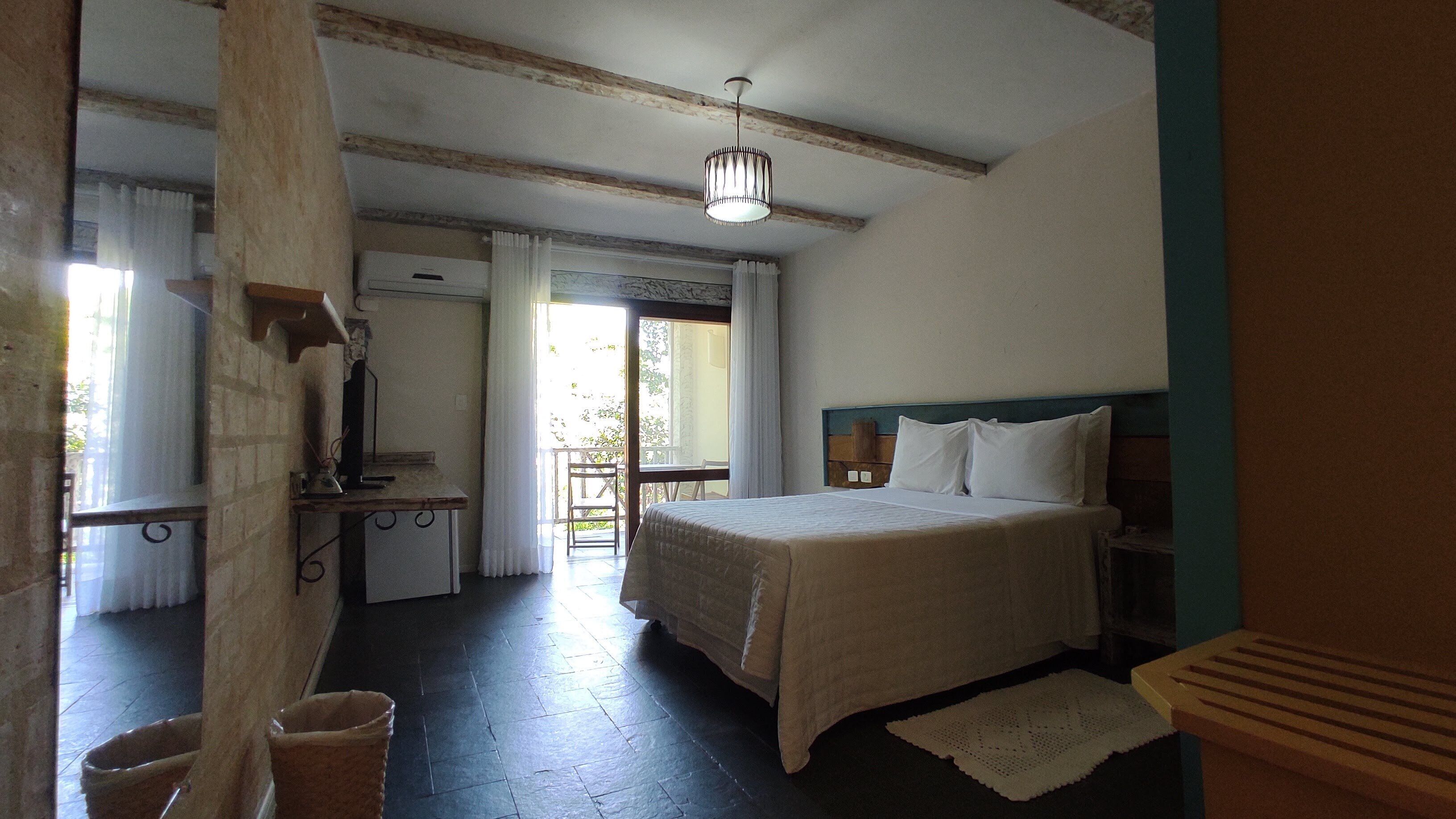 Quarto com cama, mesa de cabeceira e janela. É visível uma varanda pela janela.