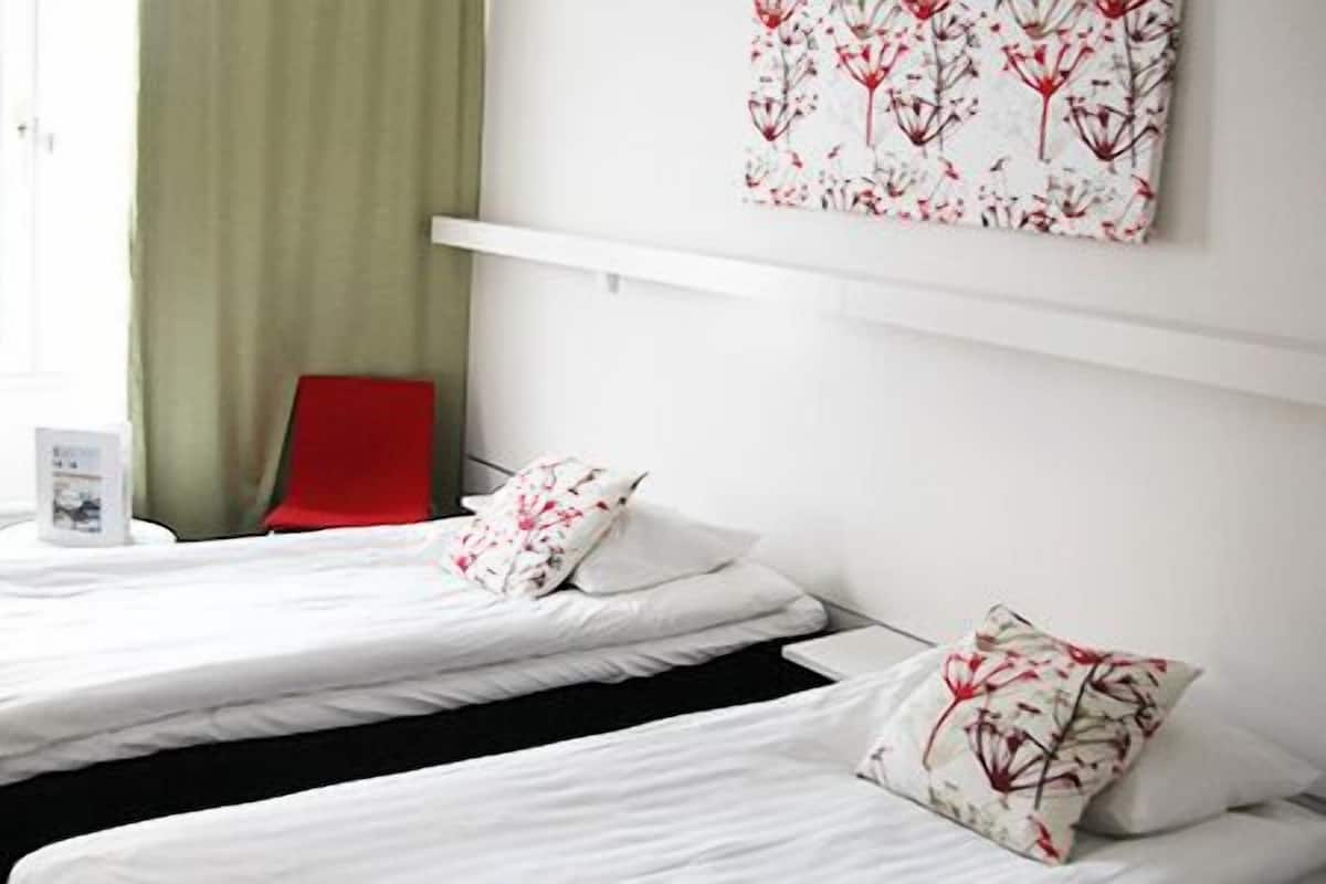 Quarto de hotel com duas camas, uma cadeira vermelha e uma prateleira na parede.