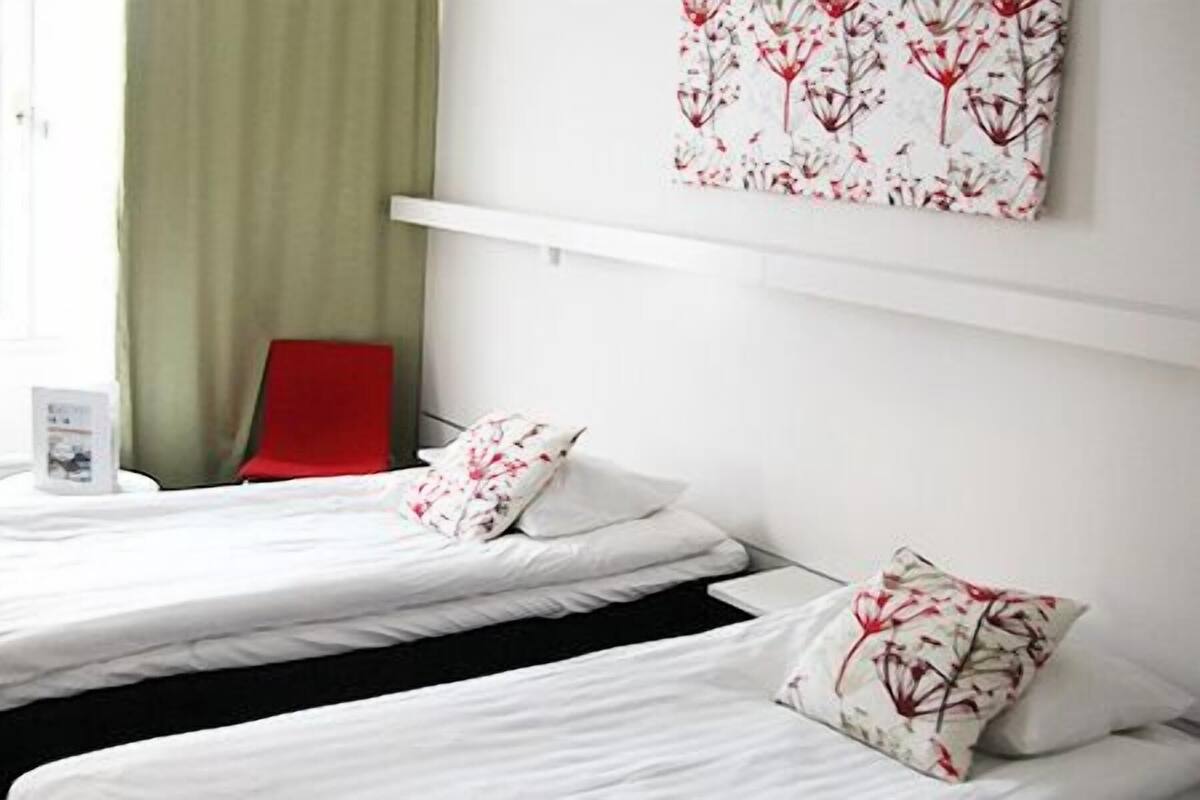 Quarto de hotel com duas camas, uma cadeira vermelha, um quadro na parede e uma janela com cortinas.