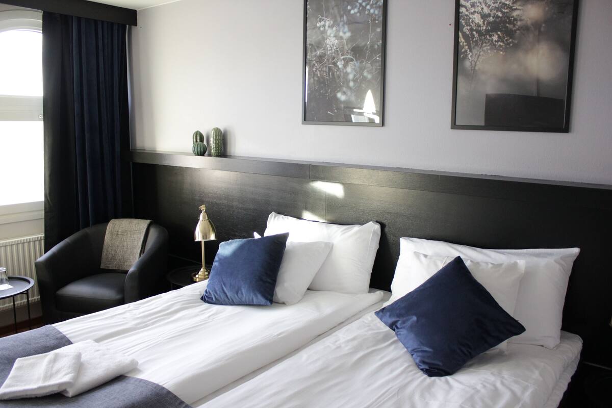 Quarto de hotel com cama, duas almofadas, uma cadeira, mesa pequena e duas quadros pendurados na parede.