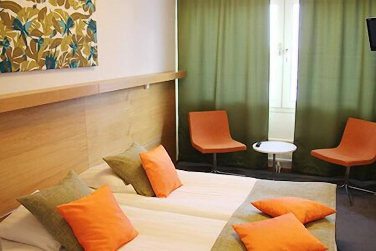 Quarto de hotel com uma cama, duas cadeiras laranja, uma mesinha e uma televisão.