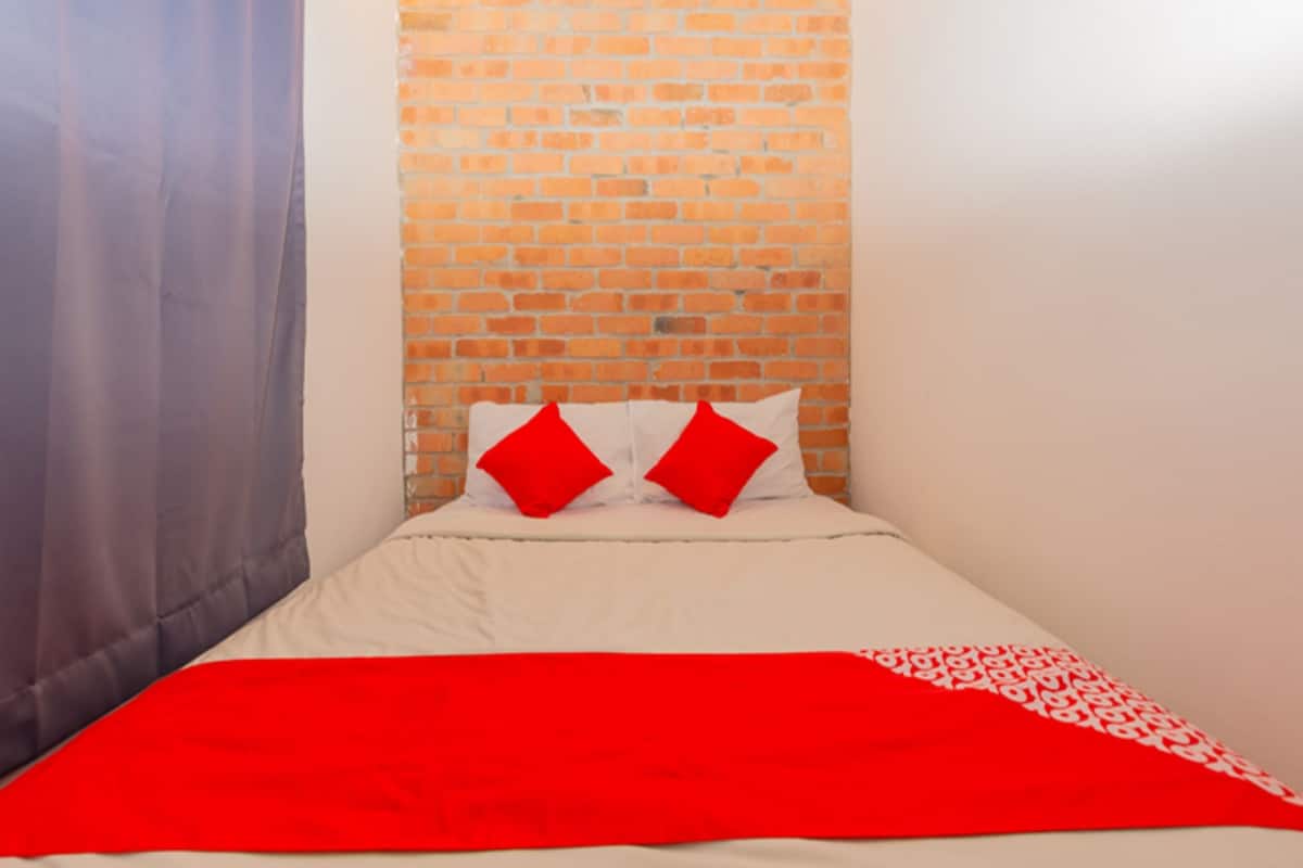 Um quarto pequeno e estreito com parede de tijolos, uma cama com roupa de cama branca e almofadas vermelhas, e uma cortina cinza de um lado.