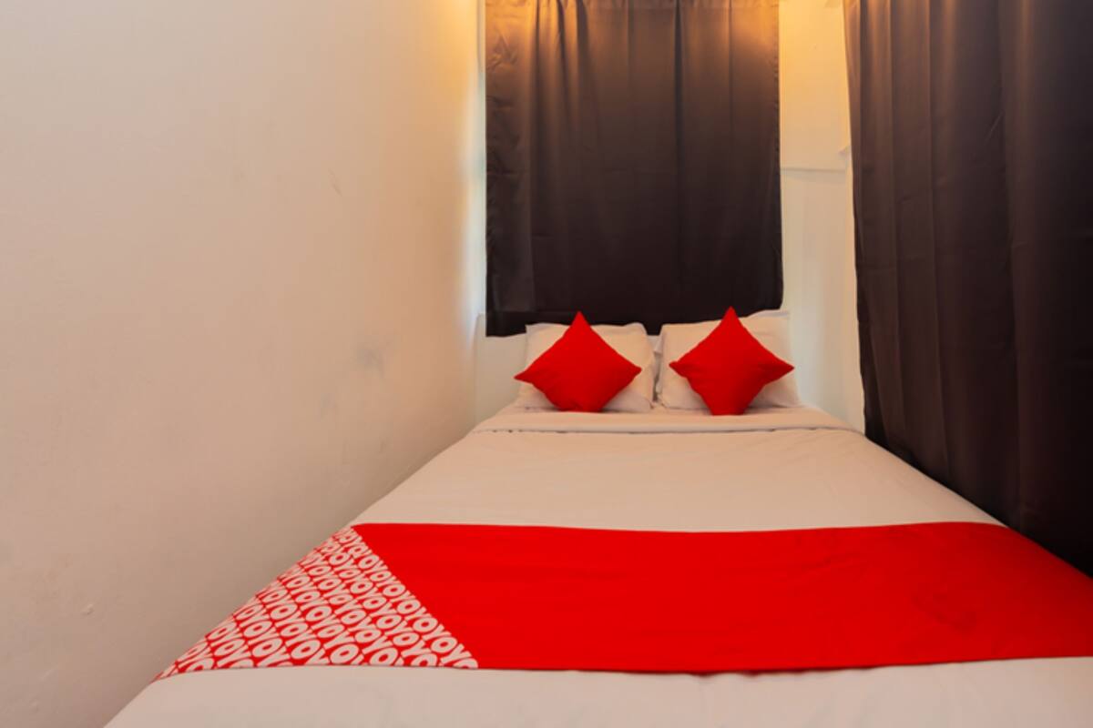 Quarto pequeno com uma cama, duas almofadas vermelhas, um cobertor com padrão vermelho e branco e uma cortina preta.