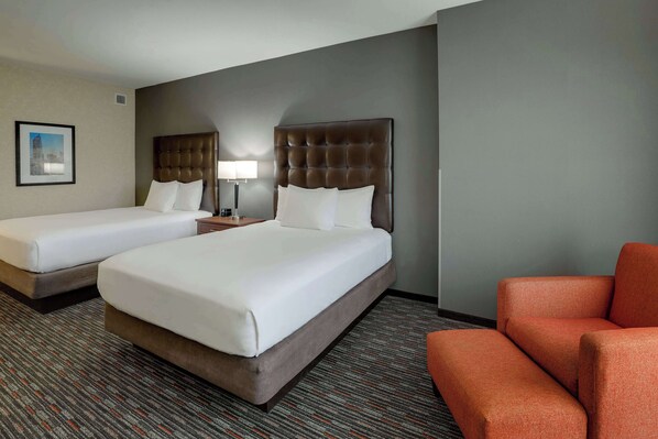 Quarto de hotel com duas camas, uma mesa de cabeceira, um abajur e uma poltrona vermelha.