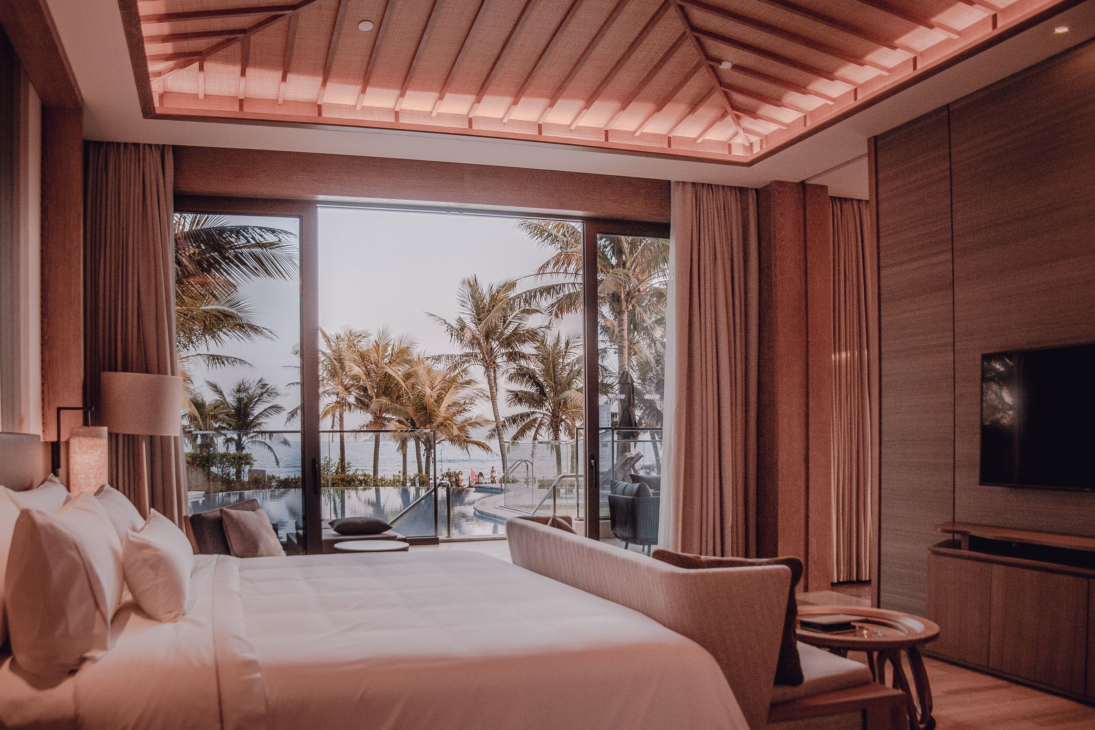 Quarto de hotel moderno com uma cama grande, TV e vista para palmeiras e uma piscina.