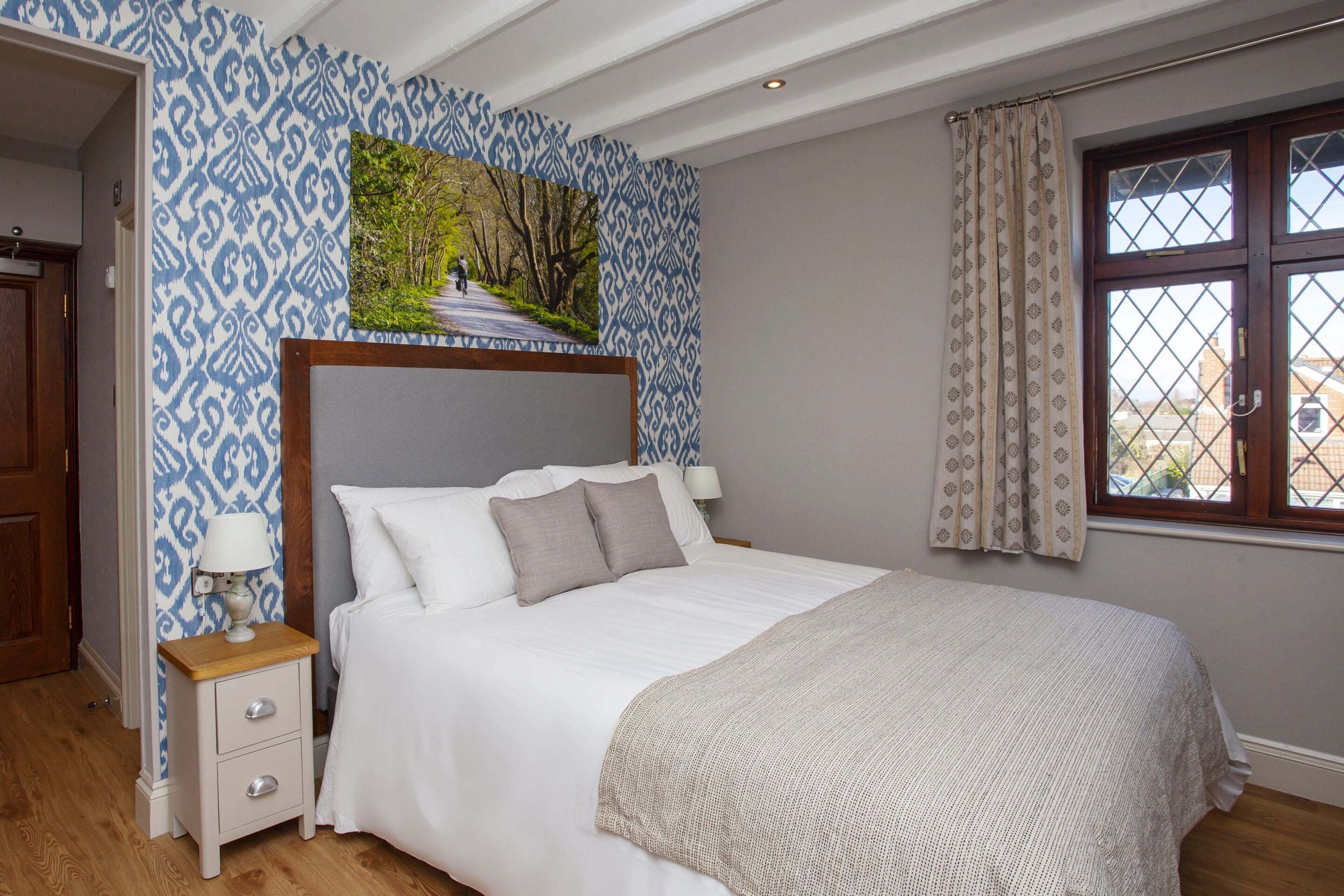 Quarto com uma cama grande, mesa de cabeceira com abajur, papel de parede estampado e janela com cortinas.