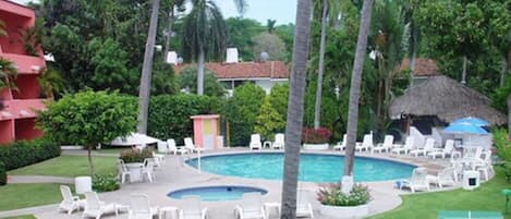 Uma área de piscina com espreguiçadeiras, palmeiras e uma estrutura com telhado de palha.