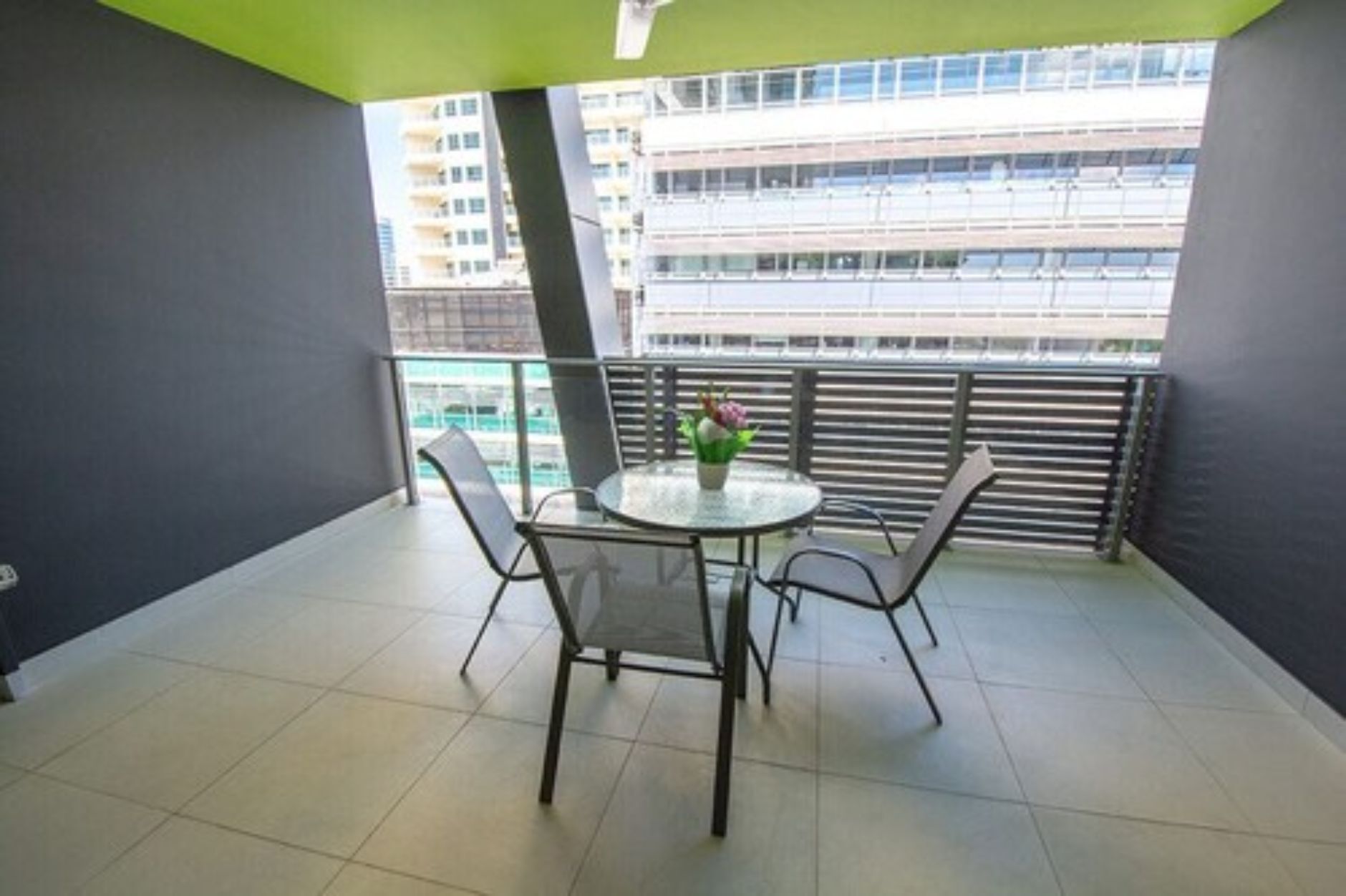 Uma varanda com uma mesa redonda e quatro cadeiras, um vaso de planta e vista para um edifício ao fundo.