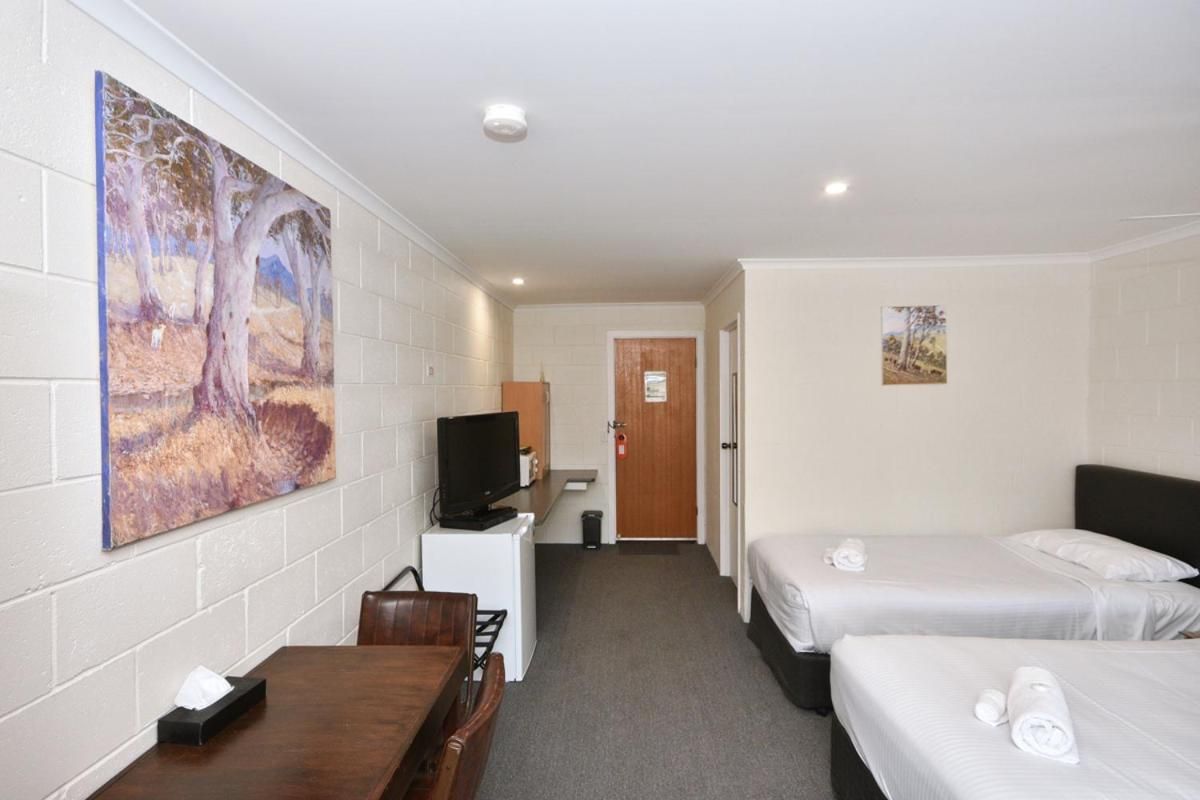 Quarto de hotel com uma cama, uma escrivaninha com televisão, um quadro na parede e uma porta que dá para outro espaço.