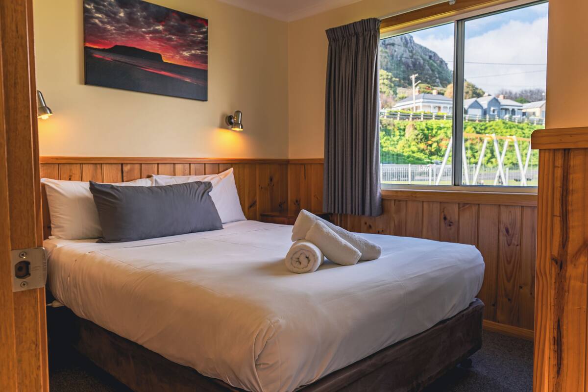 Uma cama bem arrumada com lençóis brancos e uma toalha enrolada, um quadro com uma paisagem de pôr do sol na parede, e uma janela com vista para casas e vegetação.