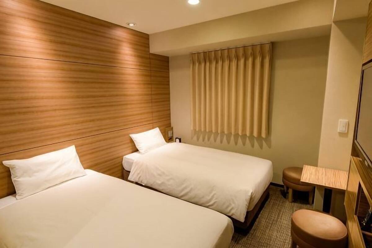 Quarto de hotel com duas camas, uma mesa pequena e janela com cortinas.
