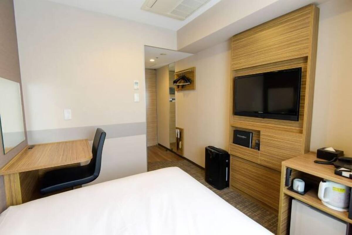 Quarto de hotel com cama, escrivaninha, cadeira, televisão, cafeteira e mini-geladeira.