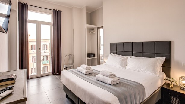 Quarto de hotel com uma cama grande, uma escrivaninha com cadeira, televisão fixada na parede e janela com cortinas.