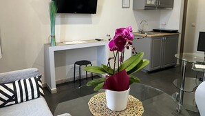 Uma sala de estar moderna com sofá, mesa de centro com um vaso de flores, televisão fixada na parede, área de cozinha com armários e pia, e um pequeno banquinho.