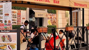 Café com mesas ao ar livre, um quadro de cardápio e uma placa indicando bar de cocktails e restaurante.