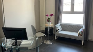 Quarto moderno com escrivaninha de vidro, computador, cadeira, sofá com almofada e janela com cortinas.