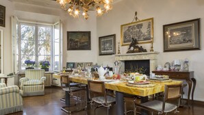 Uma sala de jantar com uma mesa de mármore, cadeiras e um candelabro. Existem quadros nas paredes e uma lareira.