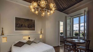 Quarto com cama grande, um candelabro, um quadro, uma mesa de jantar com cadeiras e vista pela janela.
