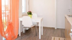 Um quarto pequeno e luminoso, com uma secretária branca, uma cadeira, um vaso de plantas pequeno e uma janela com cortinas leves.