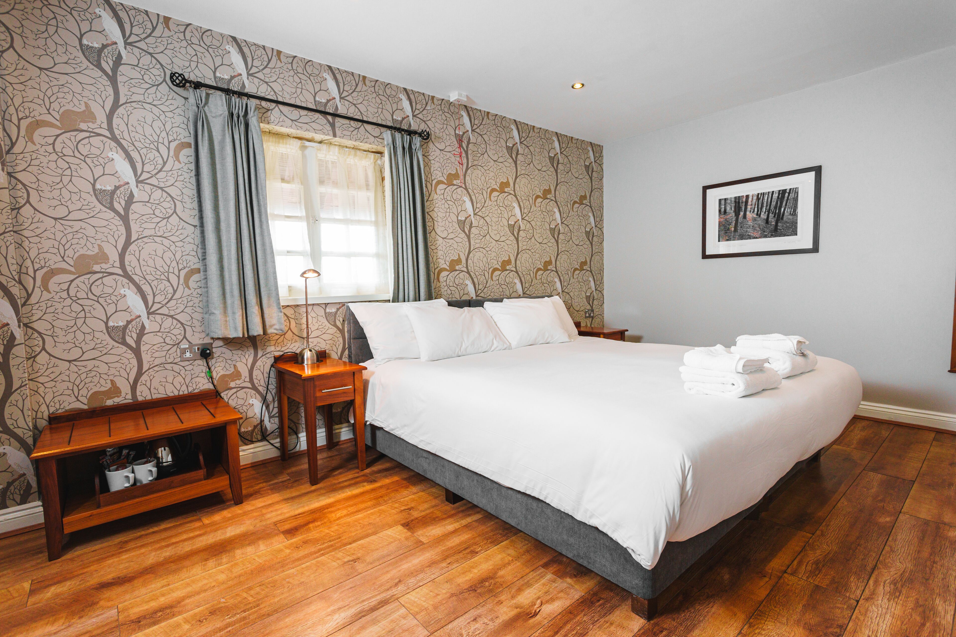 Quarto bem arrumado com cama, mesas de cabeceira, um banquinho de madeira e um quadro na parede.