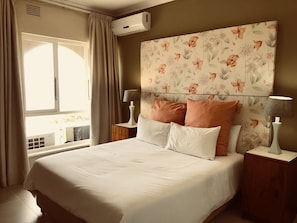 Quarto de hotel com uma cama grande, papel de parede floral, duas mesas de cabeceira com abajures e uma janela com cortinas.