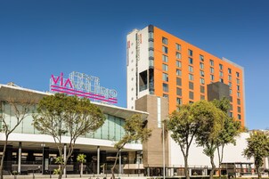 Um hotel moderno com uma placa 'VIA' em destaque, fachada de vidro e uma combinação de edifícios laranja e brancos.