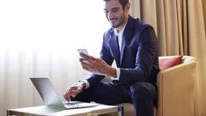 Uma pessoa de terno escuro sentada num sofá bege, com um laptop e um smartphone sobre uma mesa pequena.
