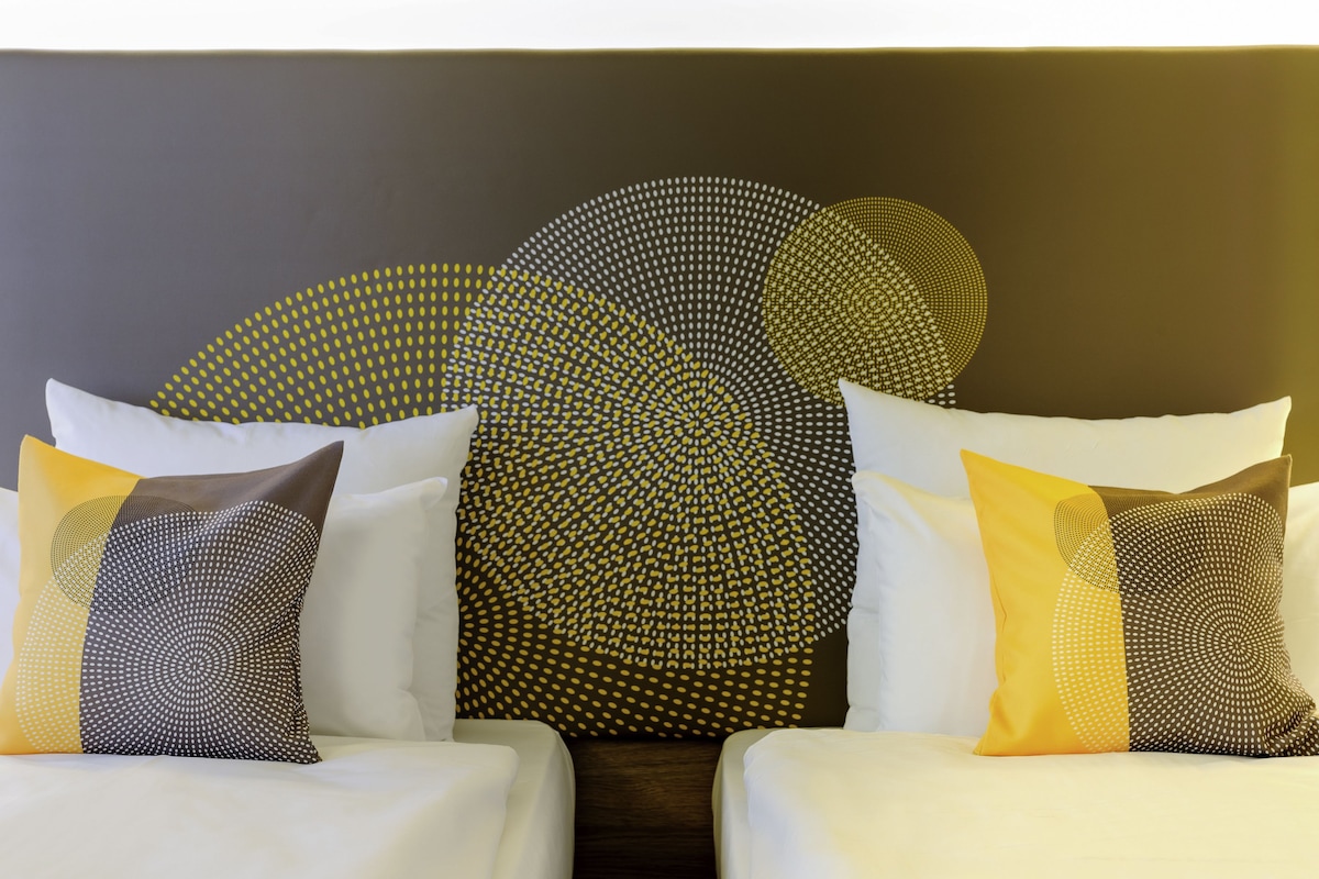 Quarto de hotel com duas camas, cada uma com um travesseiro de padrão geométrico.