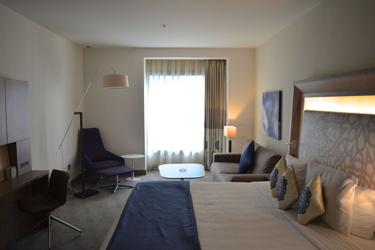 Quarto de hotel com cama, escrivaninha, cadeira, mesa pequena, luminária e televisão.