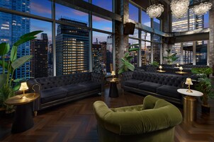 Um lounge moderno com sofás escuros, mesas redondas e amplas janelas com vista para a cidade.