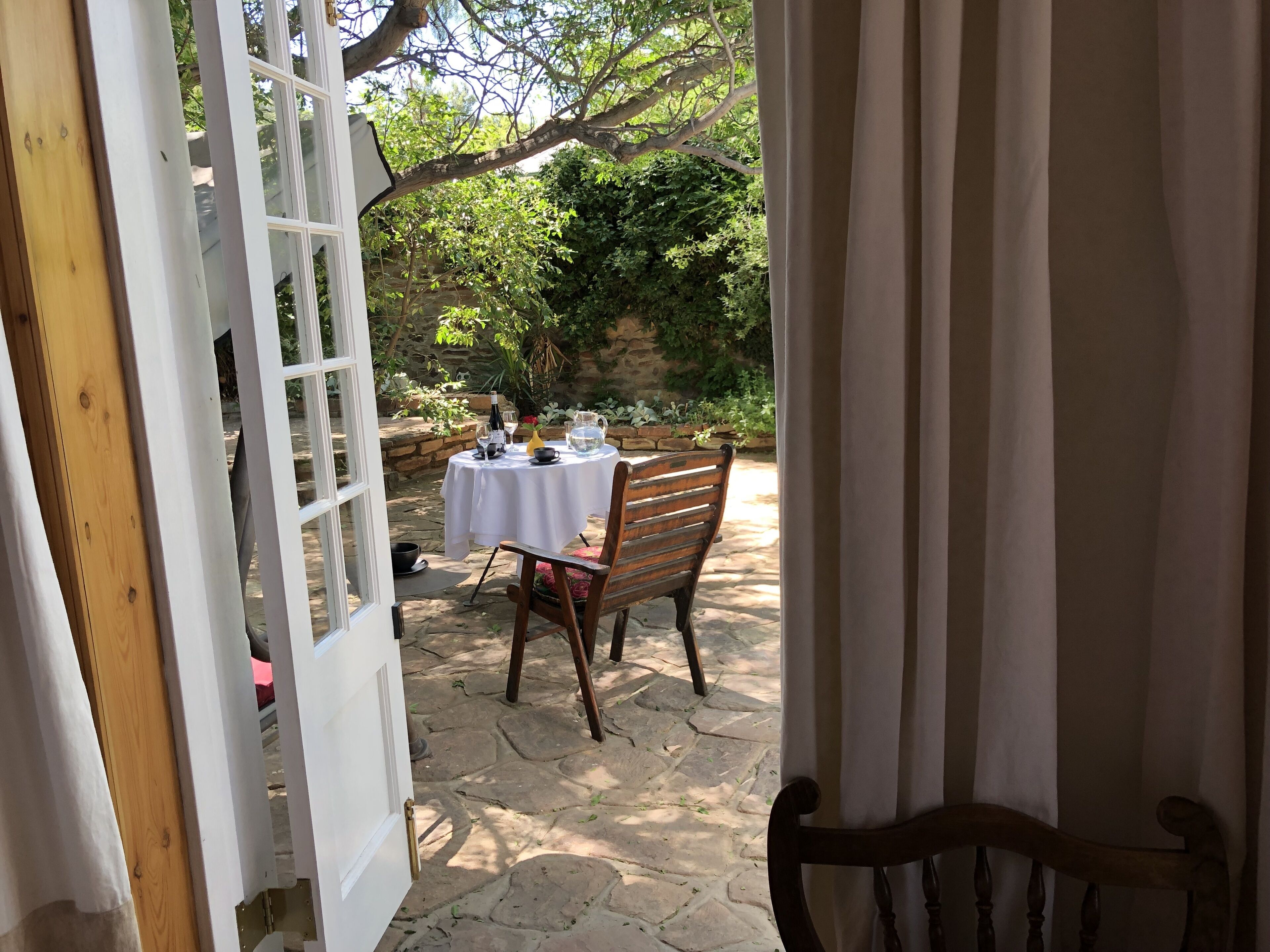 Um pátio externo com mesa redonda posta para refeições, rodeado por piso de pedra e vegetação.