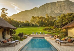 Área de piscina com espreguiçadeiras, um edifício com telhado de palha e vista para uma montanha e paisagem verde.