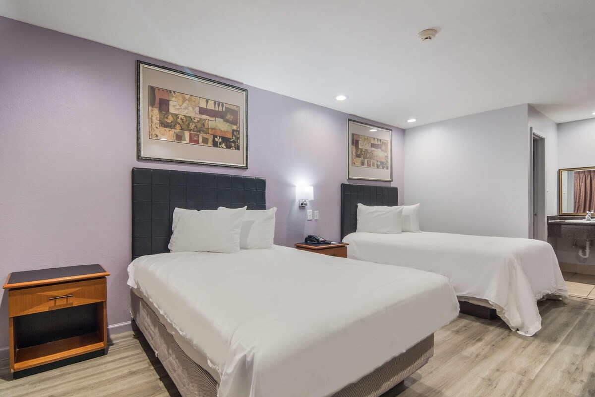 Quarto de hotel com duas camas, mesa de cabeceira, luminária e quadros nas paredes.
