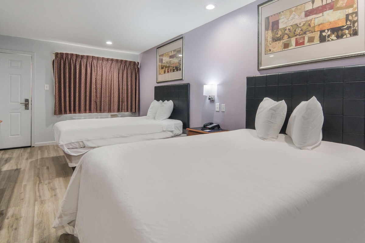 Quarto de hotel com duas camas, um quadro grande, cabeceira, mesa de cabeceira e uma porta.