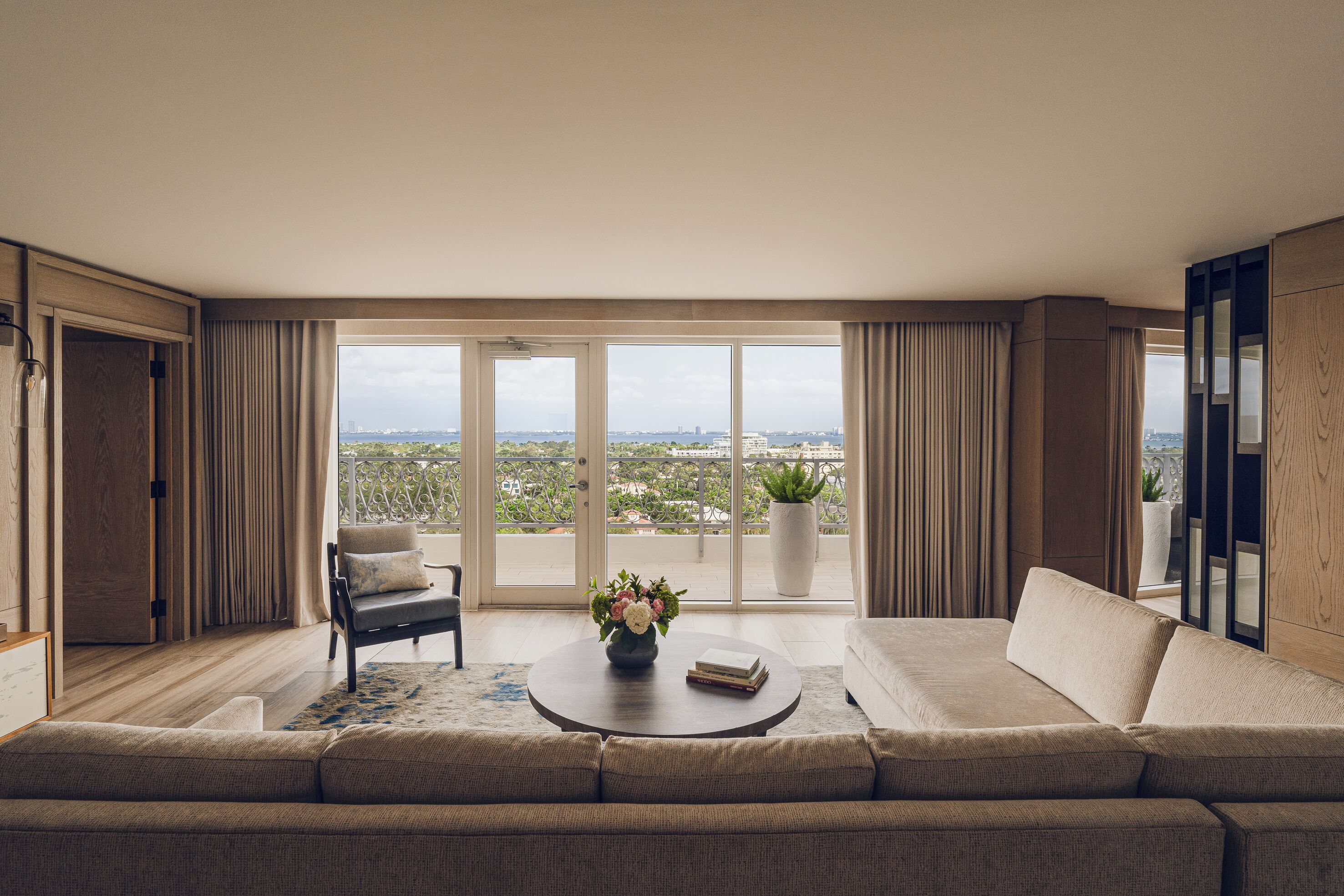 Uma sala de estar moderna com um sofá, uma mesa de centro com um livro e flores, e vista para um horizonte urbano através de grandes janelas.