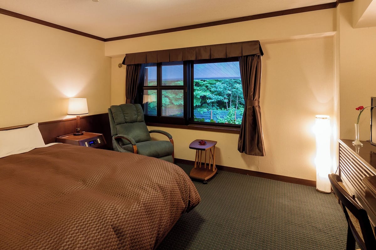 Quarto de hotel com uma cama grande, uma cadeira, uma mesa pequena, uma mesinha de cabeceira, uma janela com cortinas e uma luminária.