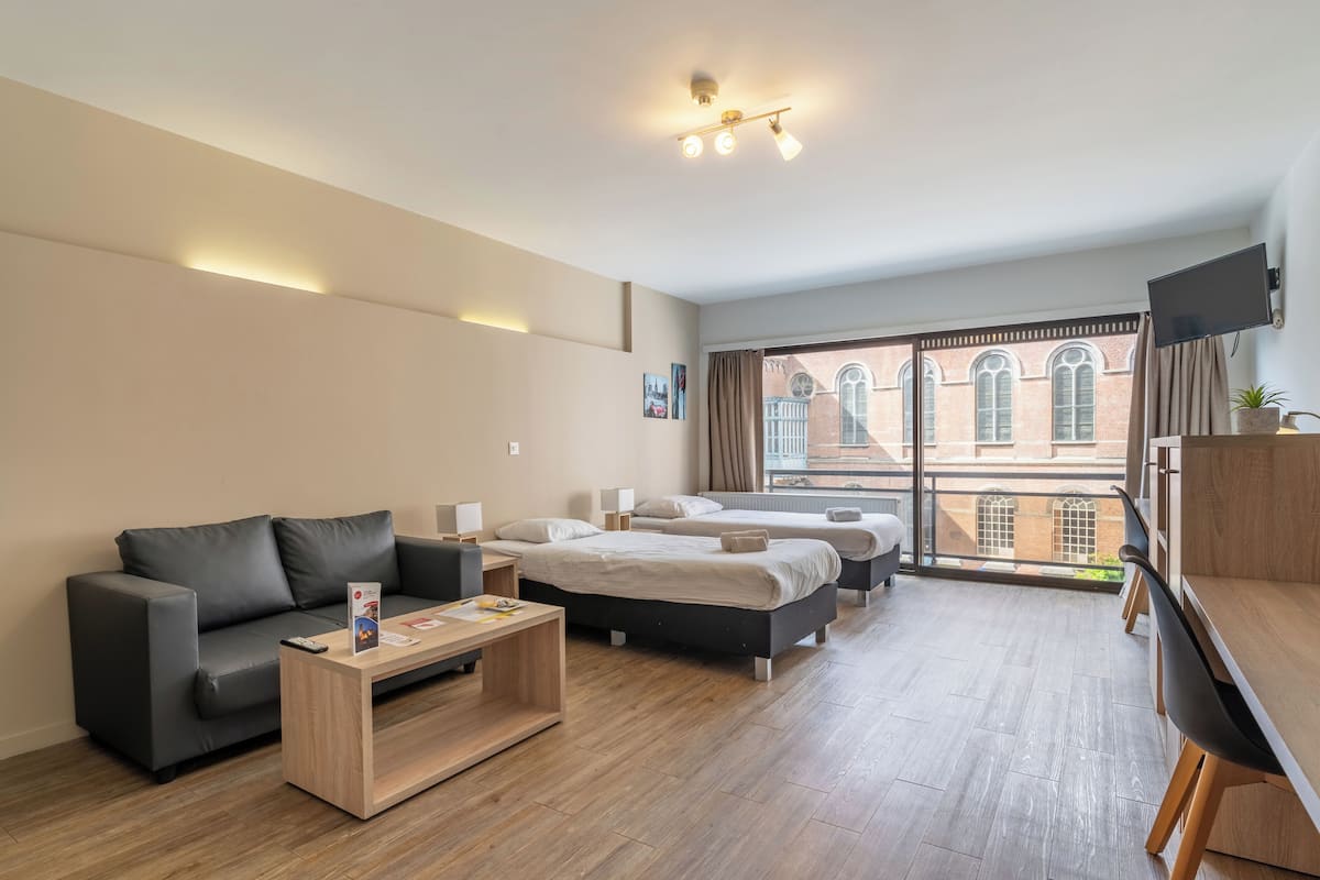 Quarto de hotel moderno com sofá, duas camas de solteiro, escrivaninha e uma grande janela com vista para prédios.