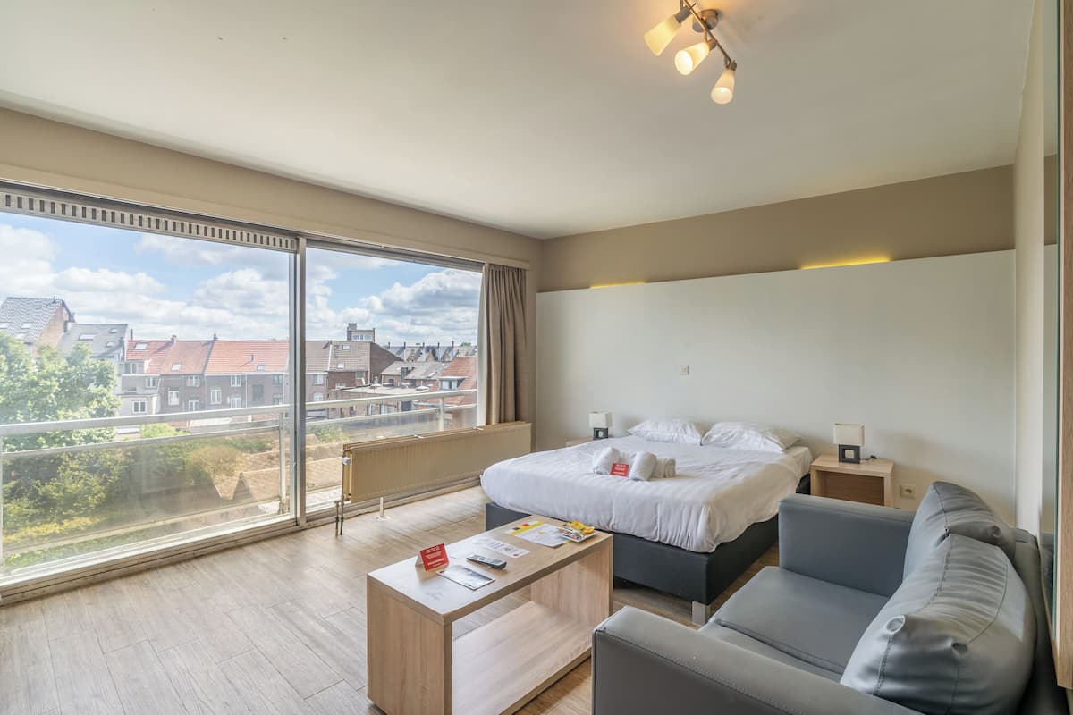Quarto de hotel moderno com uma cama grande, um sofá, uma mesa de centro de madeira e vista para a cidade através de grandes janelas.