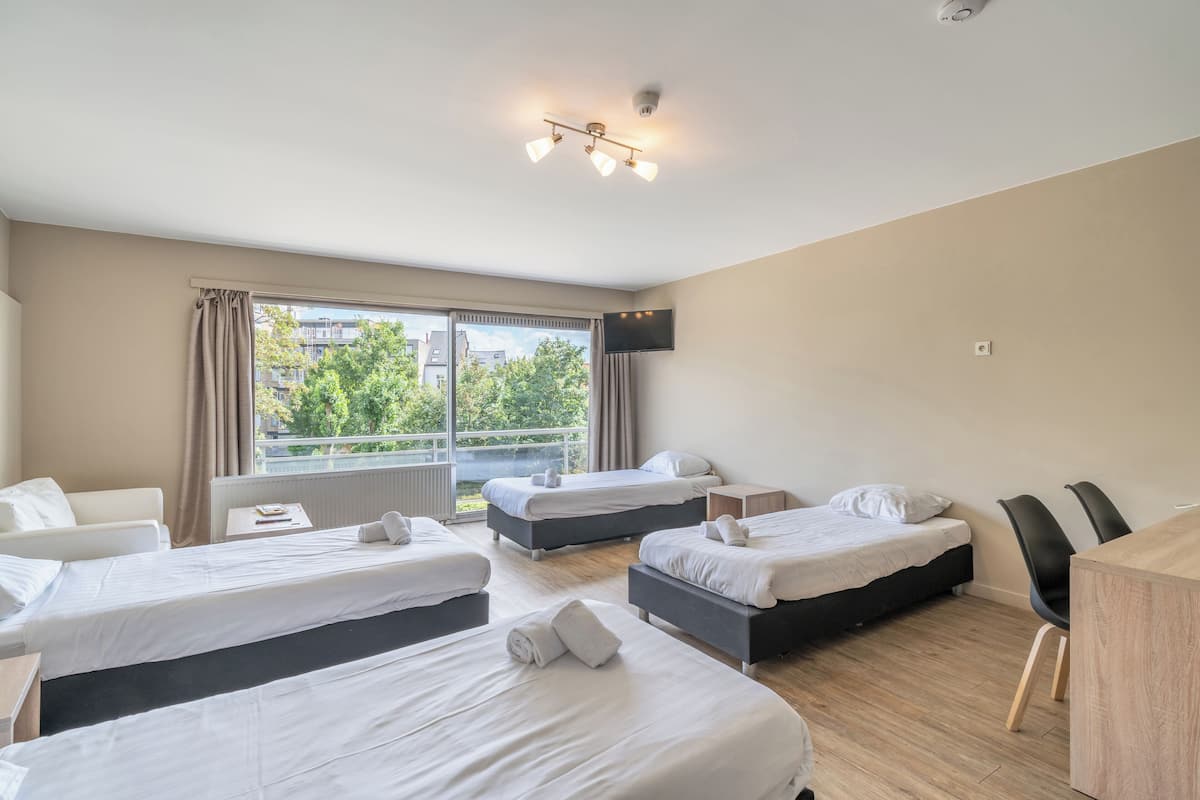 Quarto de hotel com três camas, televisão, piso de madeira e janela com vista para área verde.